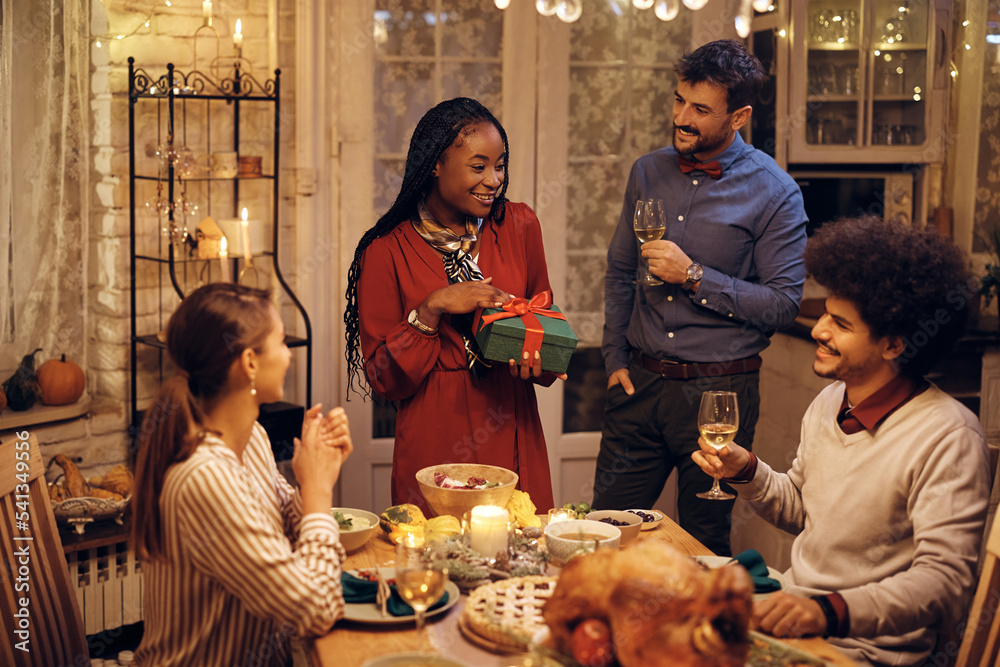 Happy African American host receiving present from her gest during dinner party on Thanksgiving ...