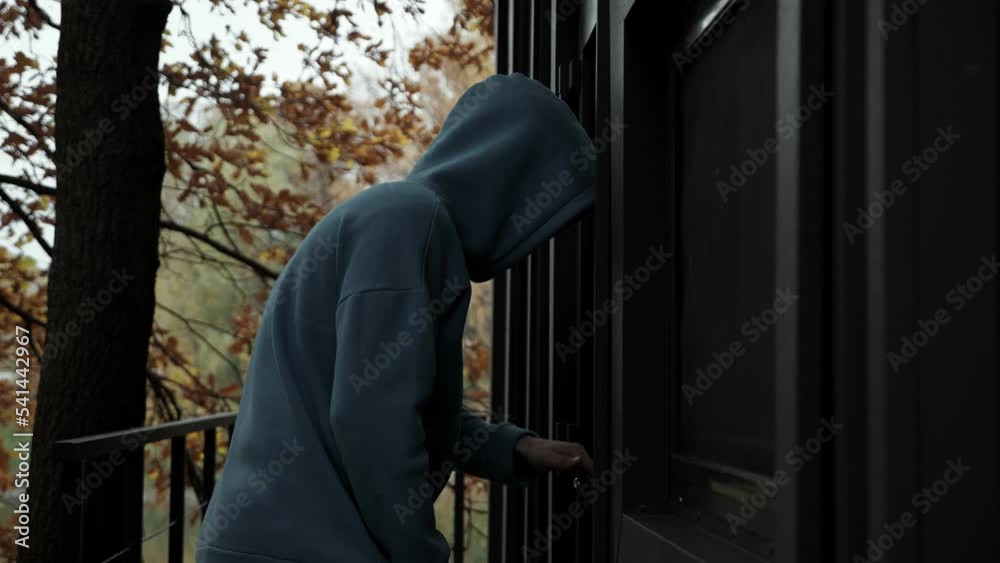 Thief in dark clothes with hood looks around and looks into house ...