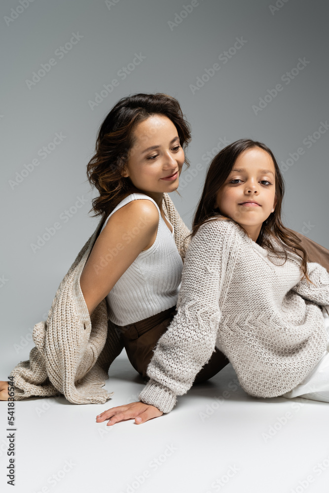 happy woman looking at trendy daughter while sitting on grey background ...
