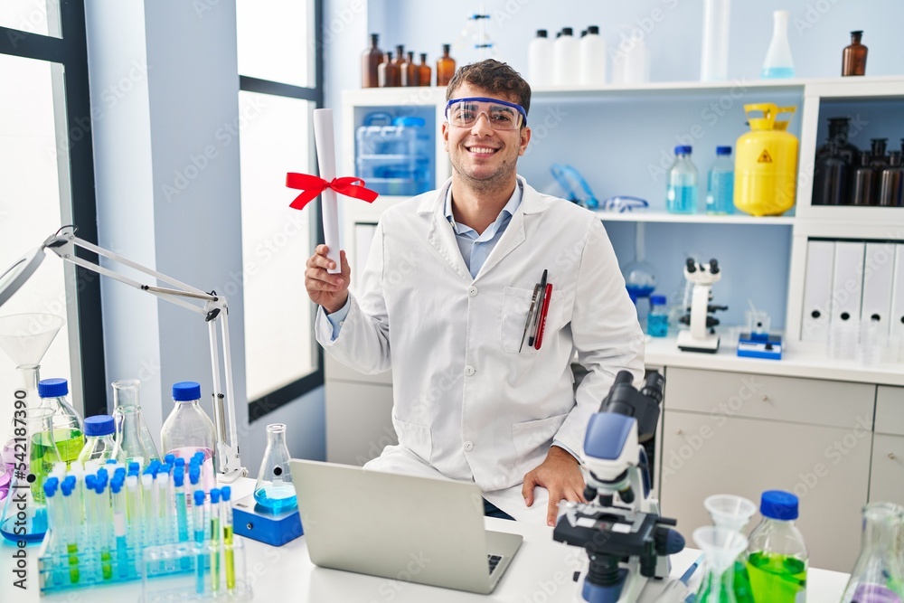 Young hispanic man working at scientist laboratory holding diploma looking positive and happy ...