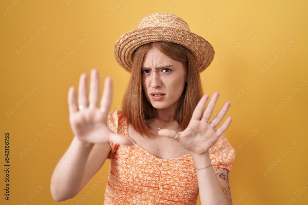 Young redhead woman standing over yellow background wearing summer hat ...