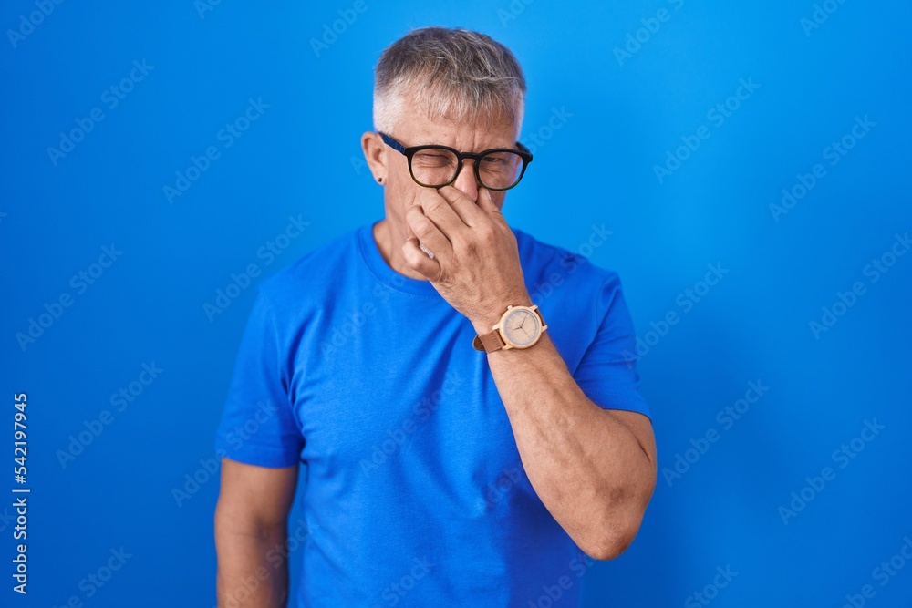Hispanic man with grey hair standing over blue background smelling ...