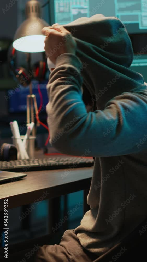 Vertical video: Masked scammer hacking computer network late at night, working on laptop and ...