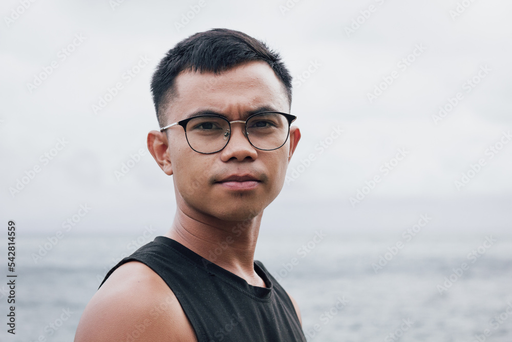 Portrait of young Filipino man with eyeglasses by the sea on cloudy day ...