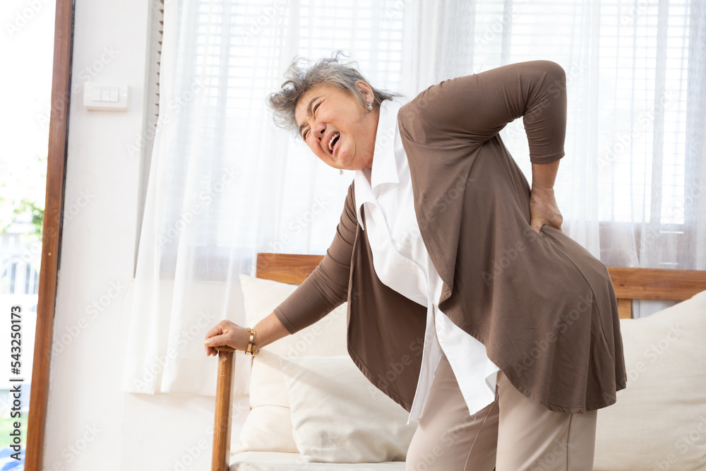 Senior Asian woman is bent over with her hand on the couch because she ...
