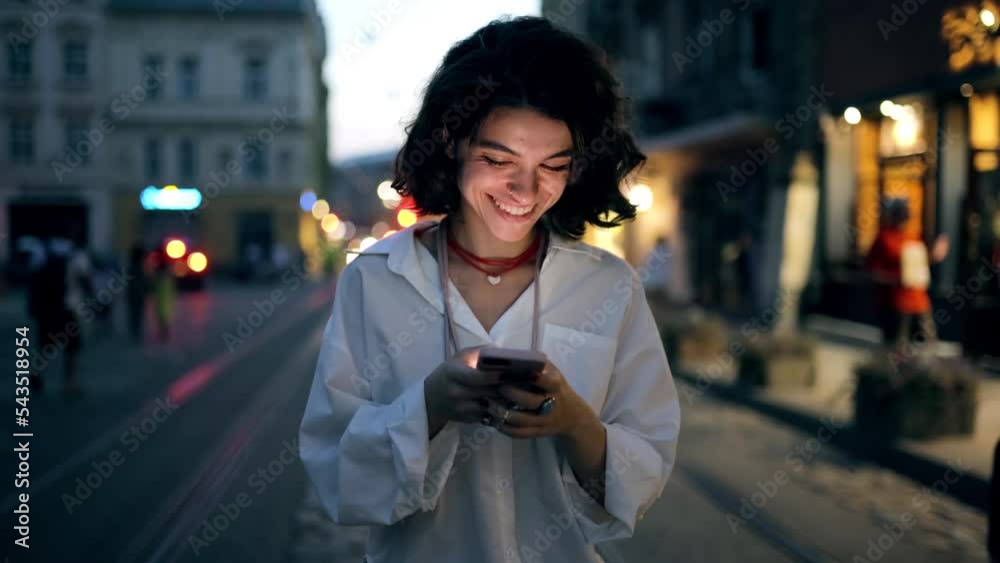 Smiling woman using smartphone on street with night city lights on ...
