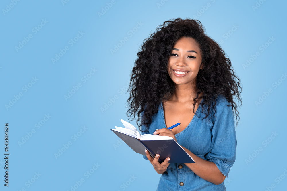 Young black woman taking notes and smiling, copy space blue background ...