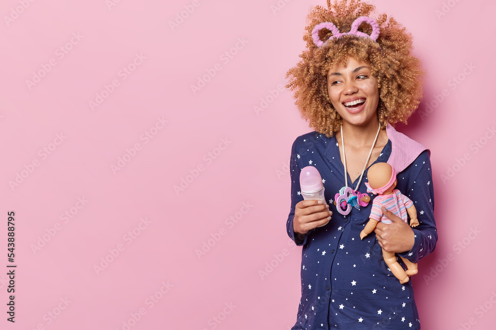 Positive future mother with tummy holds feeding bottle of milk and doll ...