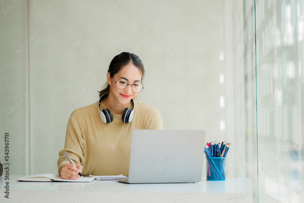 Asian schoolgirl studying and learning online video call using laptop wearing headphones online ...