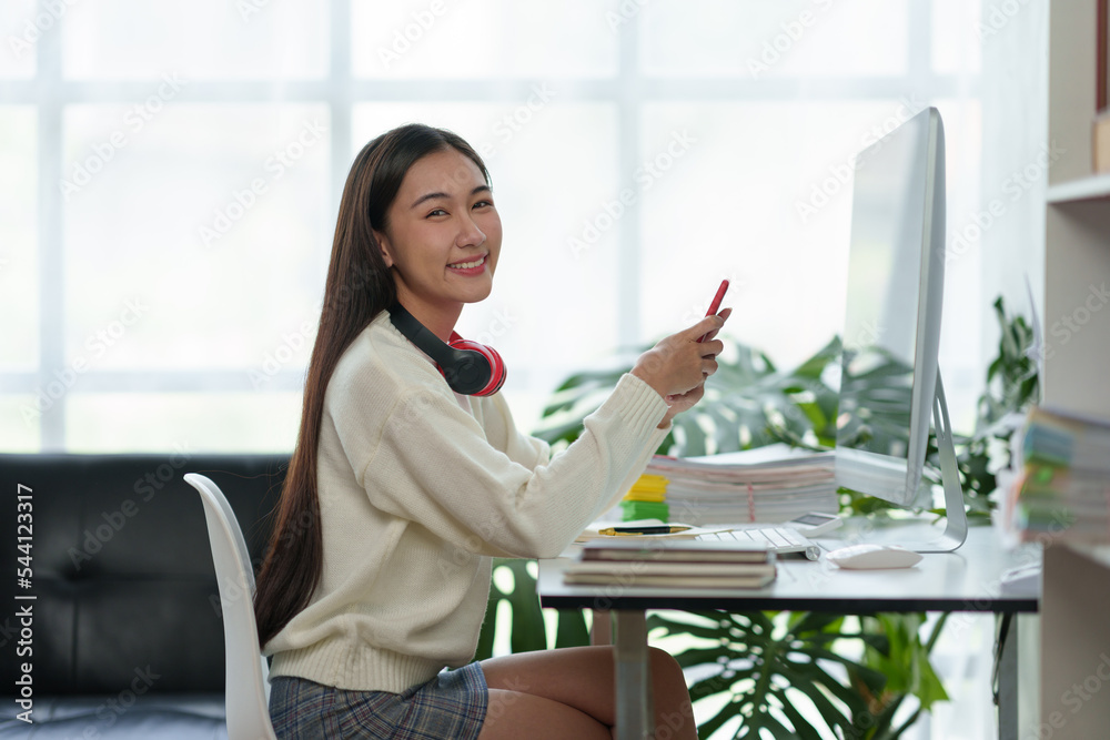 Smiling and charming Asian woman sit at her desk with a smartphone ...