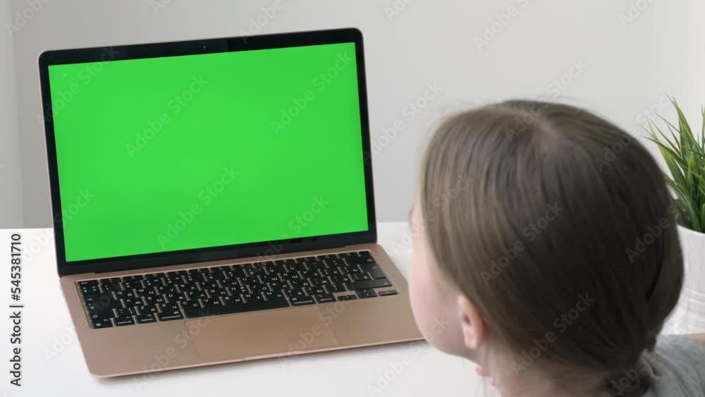 Schoolgirl studying during online lessons at home in front of green screen laptop. Chroma key, e ...