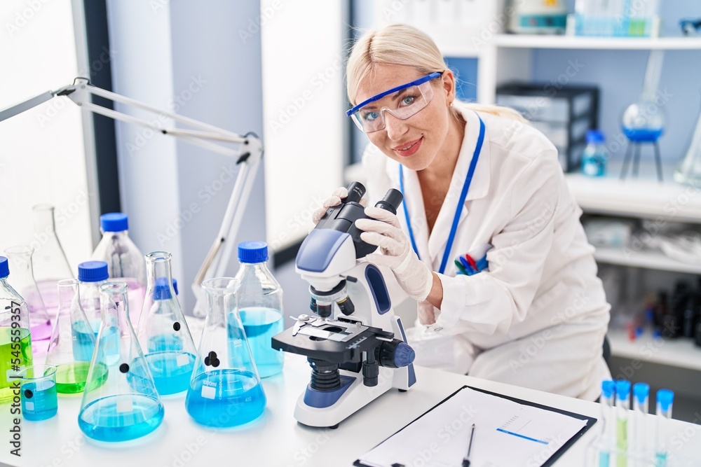Young blonde woman wearing scientist uniform using microscope at ...