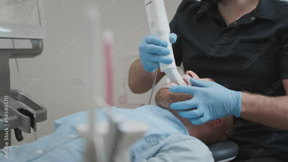 A male orthodontist doctor scans a man's teeth and jaw in the clinic's ...