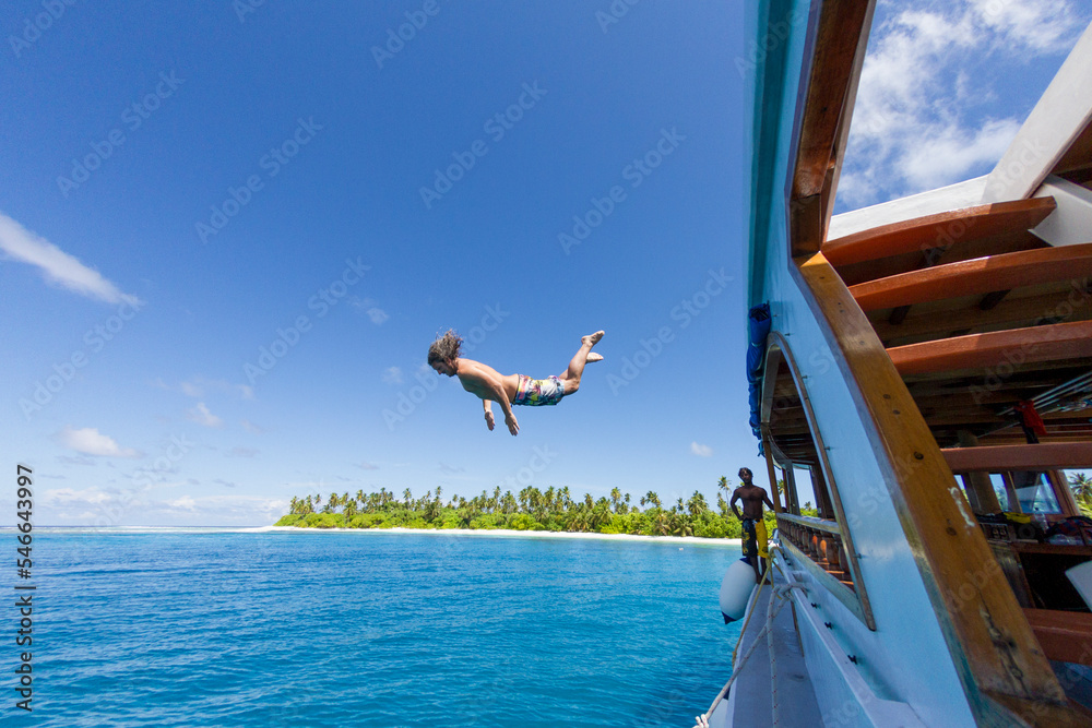 Young man jumping from the deck of a boat with an atoll full of palm ...