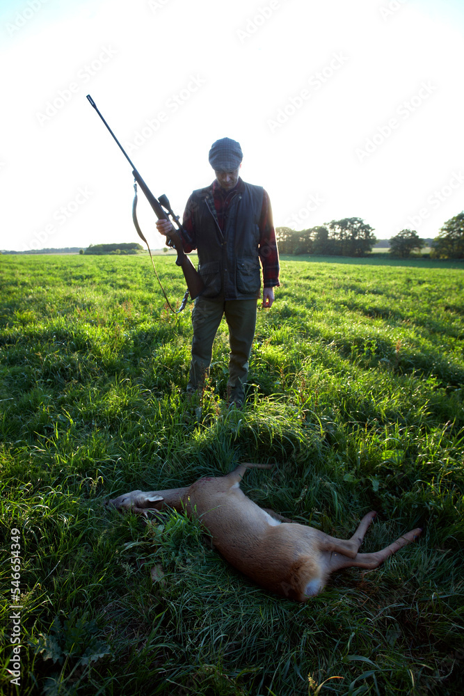 Fotka „A hunter stands over his kill in a grassy field, during a deer ...