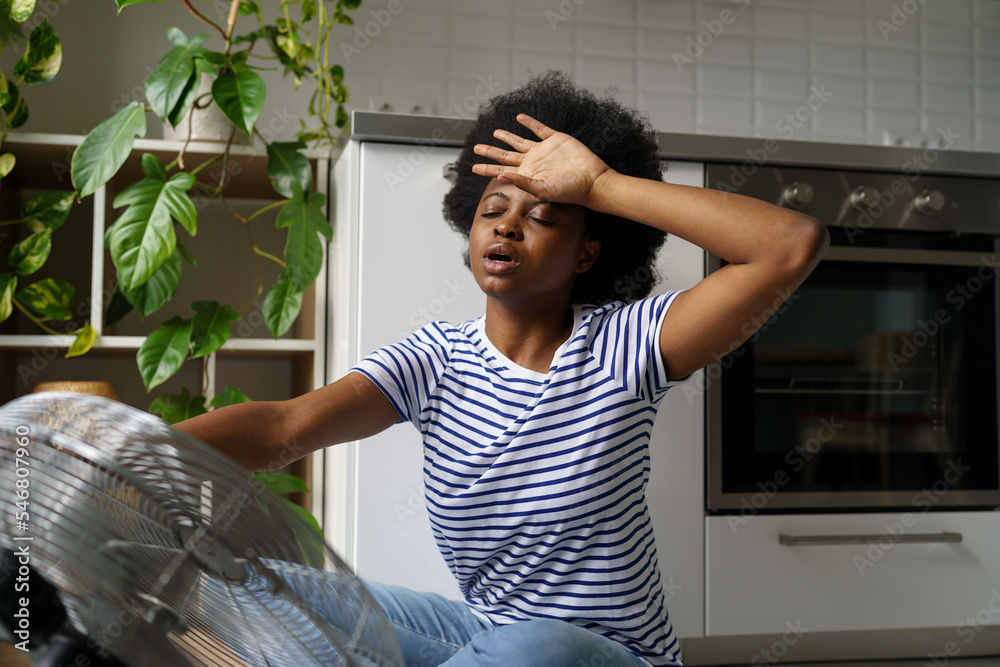 Sweaty overheated young African woman using electric fan ventilator ...