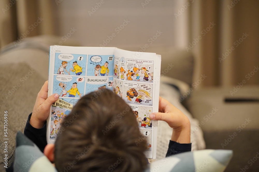 Child reading a Polish Ptys I Bill comic book on a sofa in a living ...