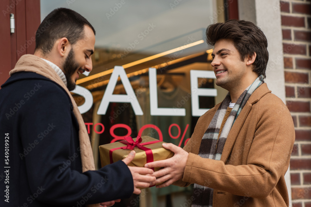 happy and stylish gay couple holding gift box near showcase with sale ...