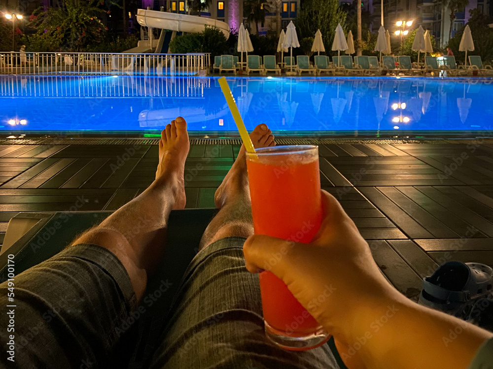 Men's legs on a deck chair by the pool at night, relaxing with an ...
