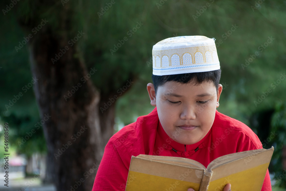 Asian muslim boy in red suit sits in school park and reading his daily ...