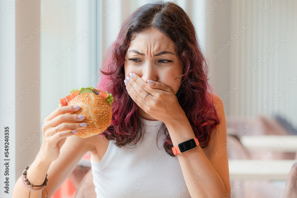 A girl eats a burger in a fast food cafe and feels nausea and heartburn ...