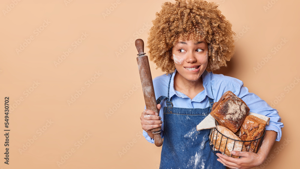 Busy female baker bites lips looks aside holds wooden rolling pin and ...