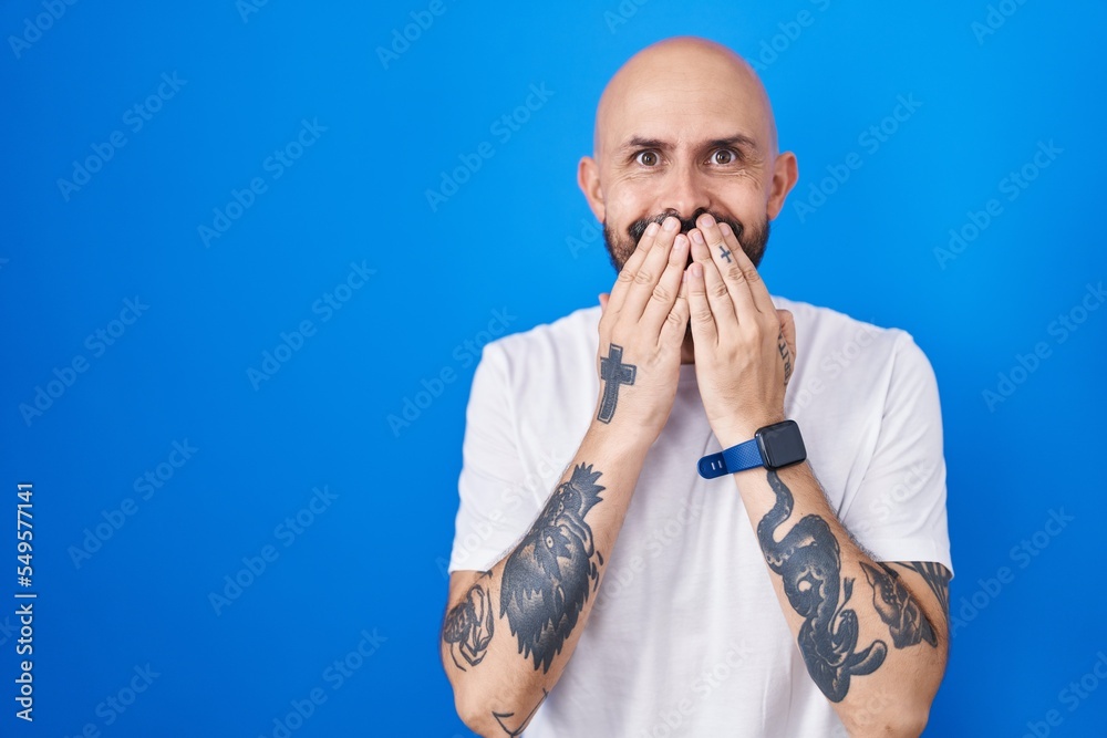 Hispanic man with tattoos standing over blue background laughing and ...