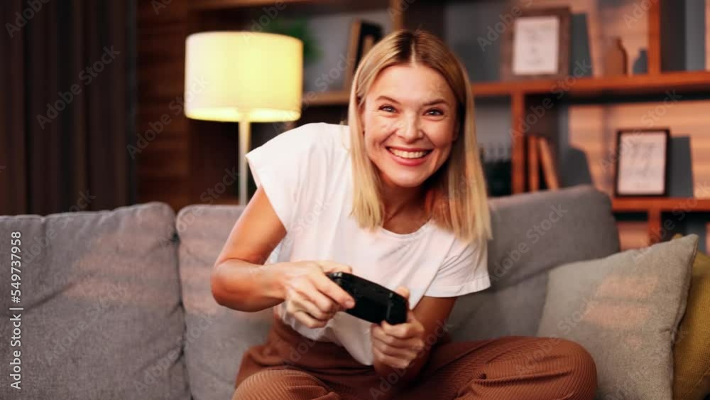 Excited middle aged blond woman gamer sitting on a couch playing in ...