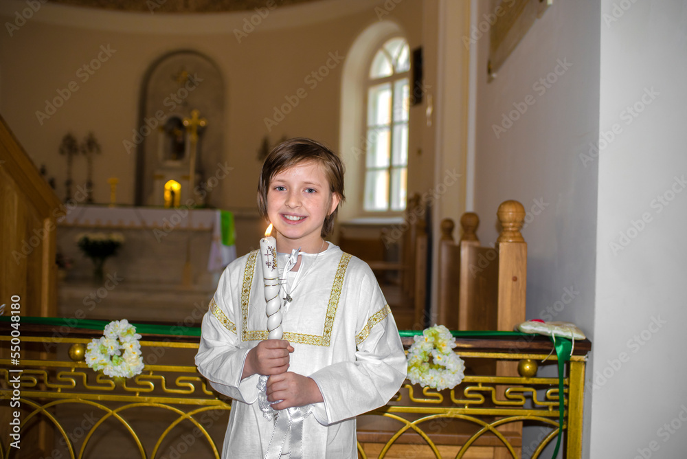 a handsome smiling boy in white clothes with a church candle stands in ...