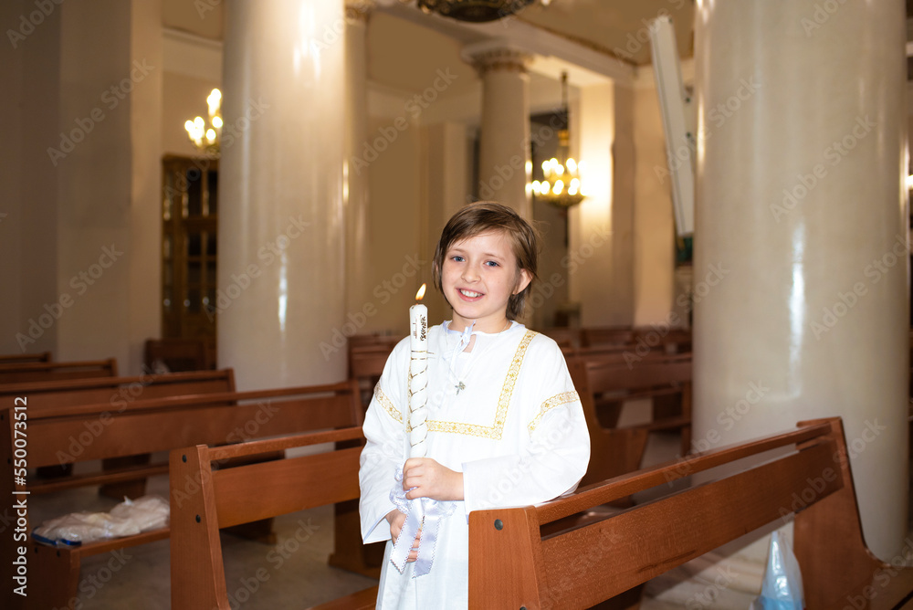a handsome smiling boy in white clothes with a church candle stands in ...