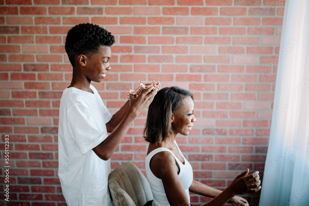 Preteen black boy placing tiara on smiling seated mom Stock-Foto ...