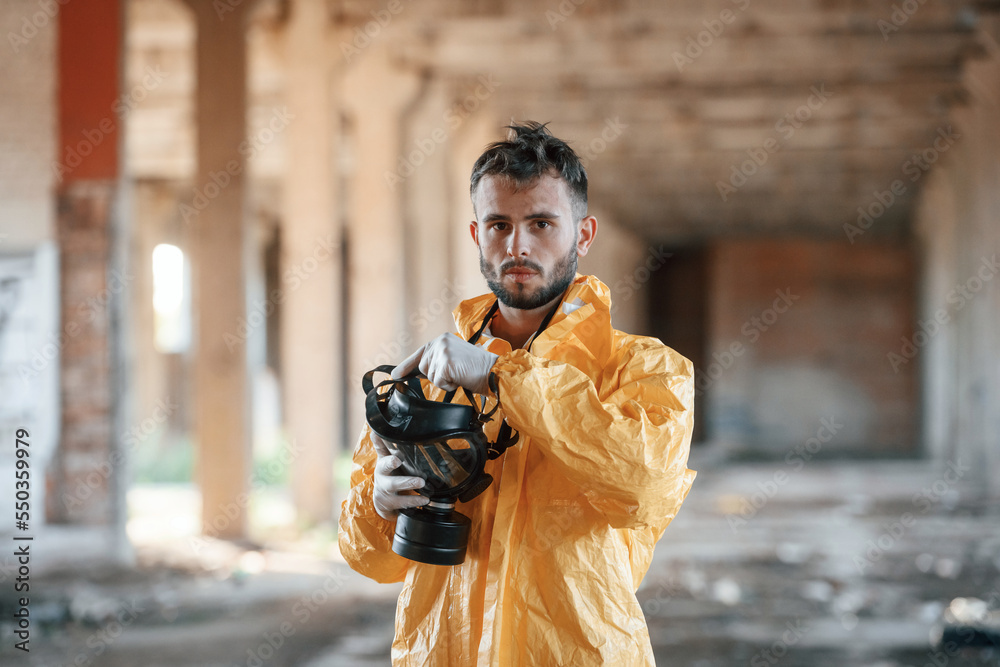 Holding gas mask. Man dressed in chemical protection suit in the ruins ...