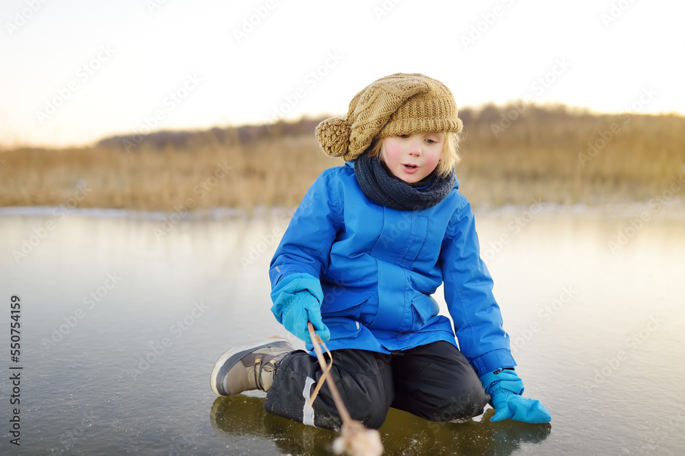 Cute preschool boy is playing on the ice of a frozen lake or river on a ...