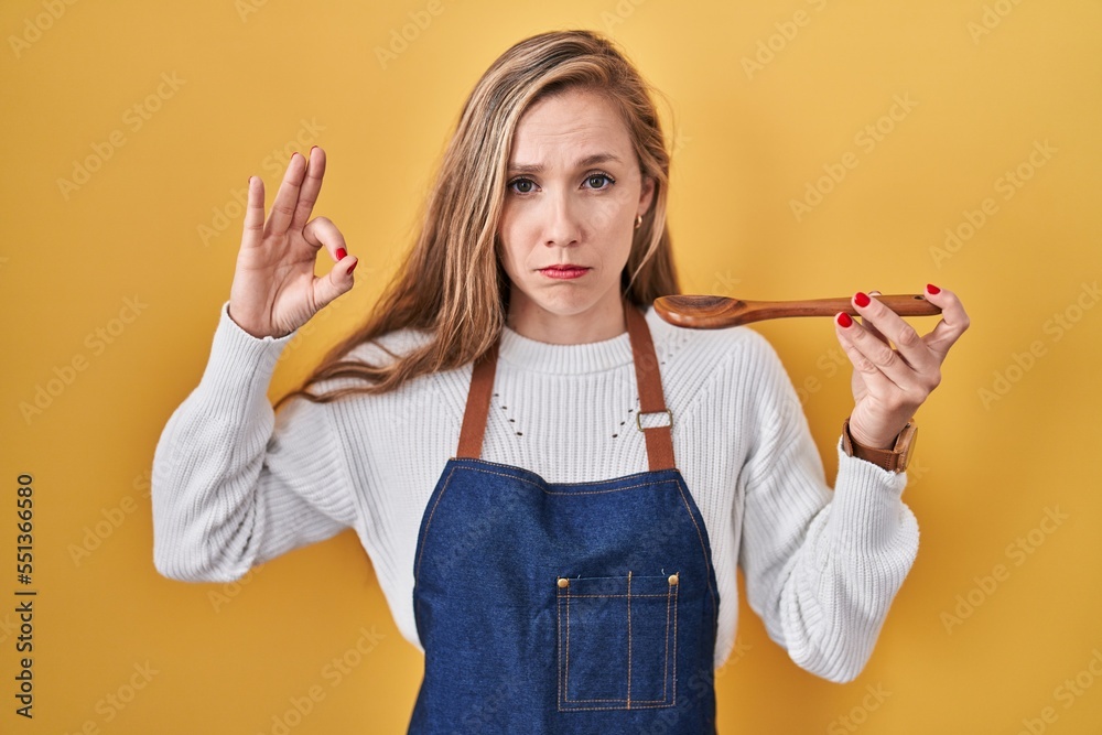 Young blonde woman wearing apron tasting food holding wooden spoon ...