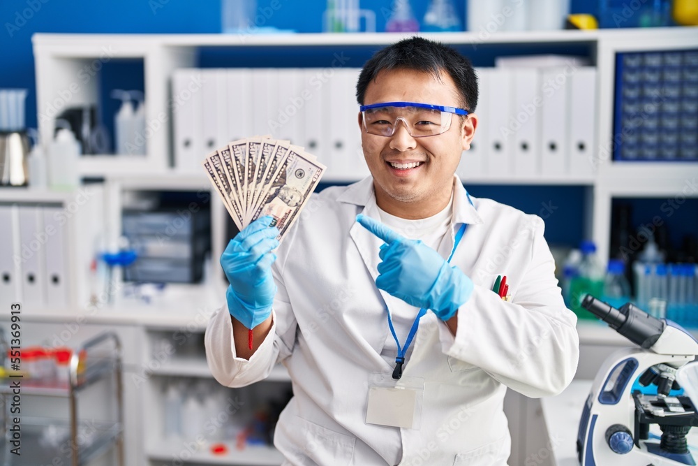 Young chinese man working at scientist laboratory holding money smiling ...