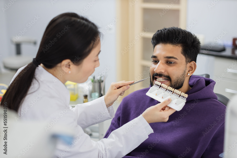 Asian dentist checking and selecting color of young bearded man teeth ...