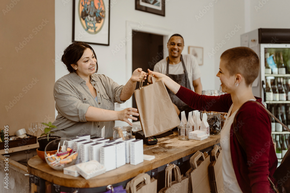 Happy female owner giving paper bag to customer at checkout counter in ...
