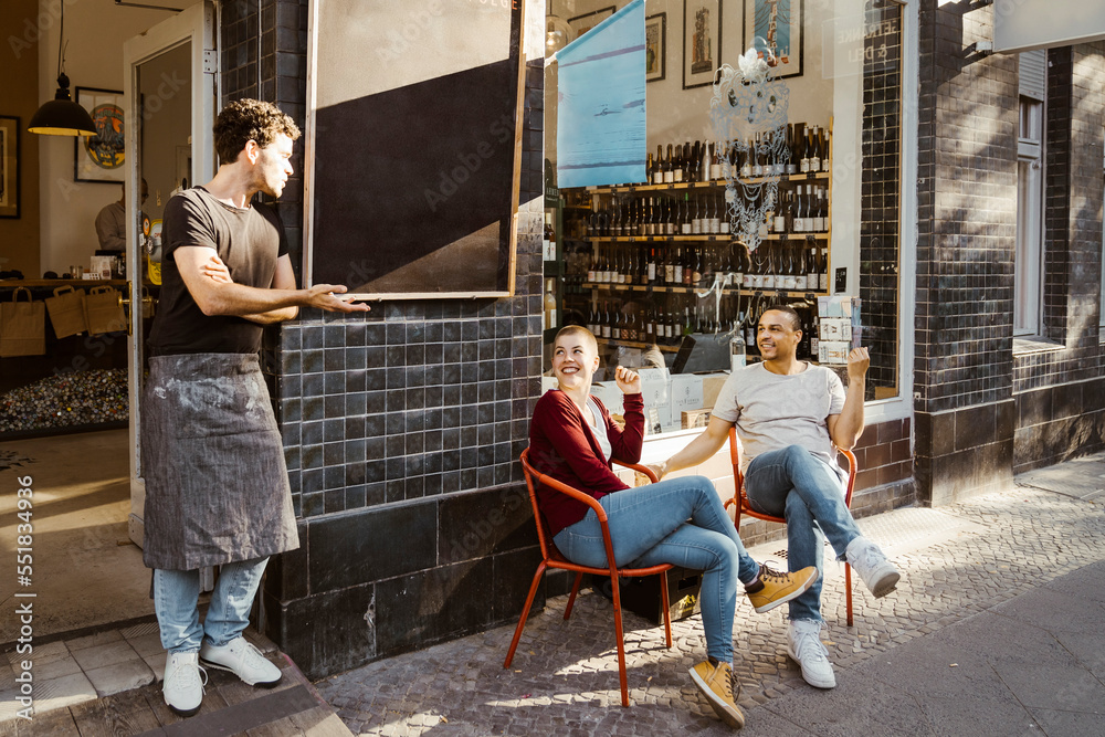 Male owner talking to customers sitting on chair at sidewalk outside ...