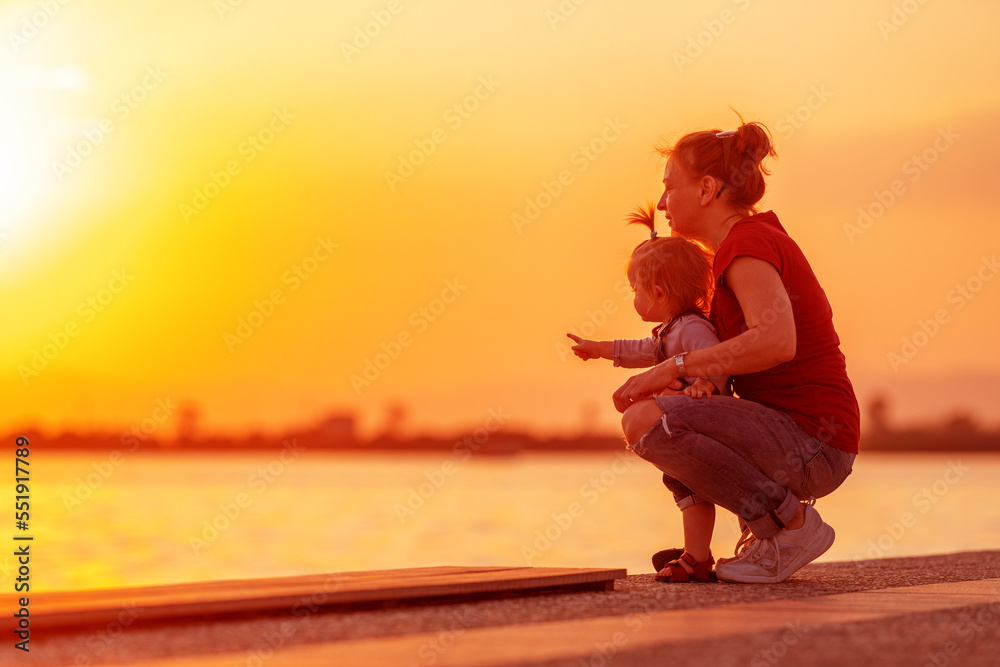 Mom and daughter are sitting on the embankment at sunset. The family is ...