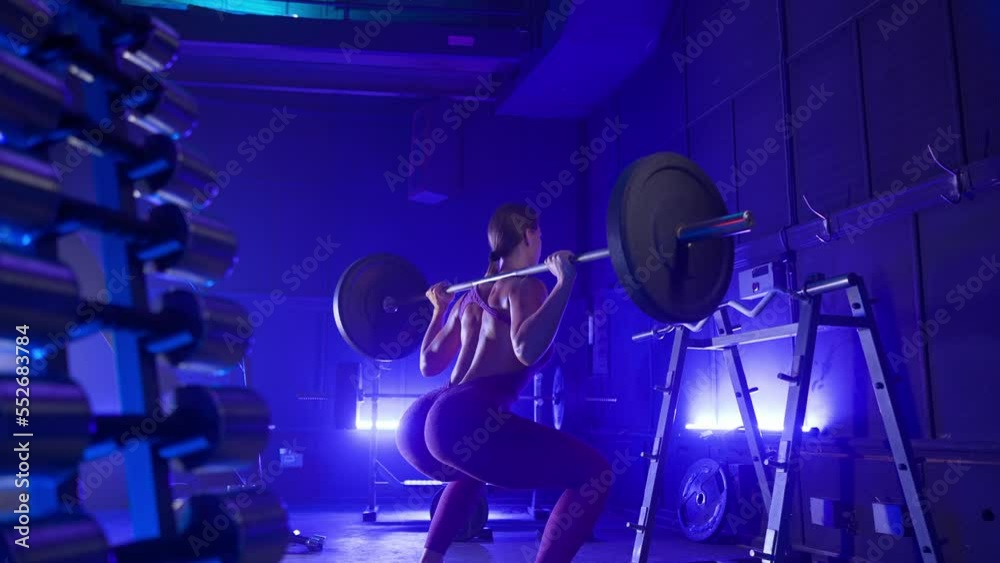 Back view of sporty fitness woman squatting with barbell in dark gym ...