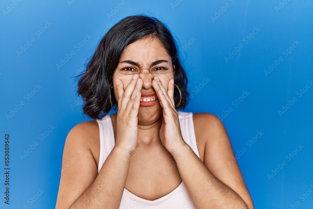 Young hispanic woman standing over blue background smelling something ...