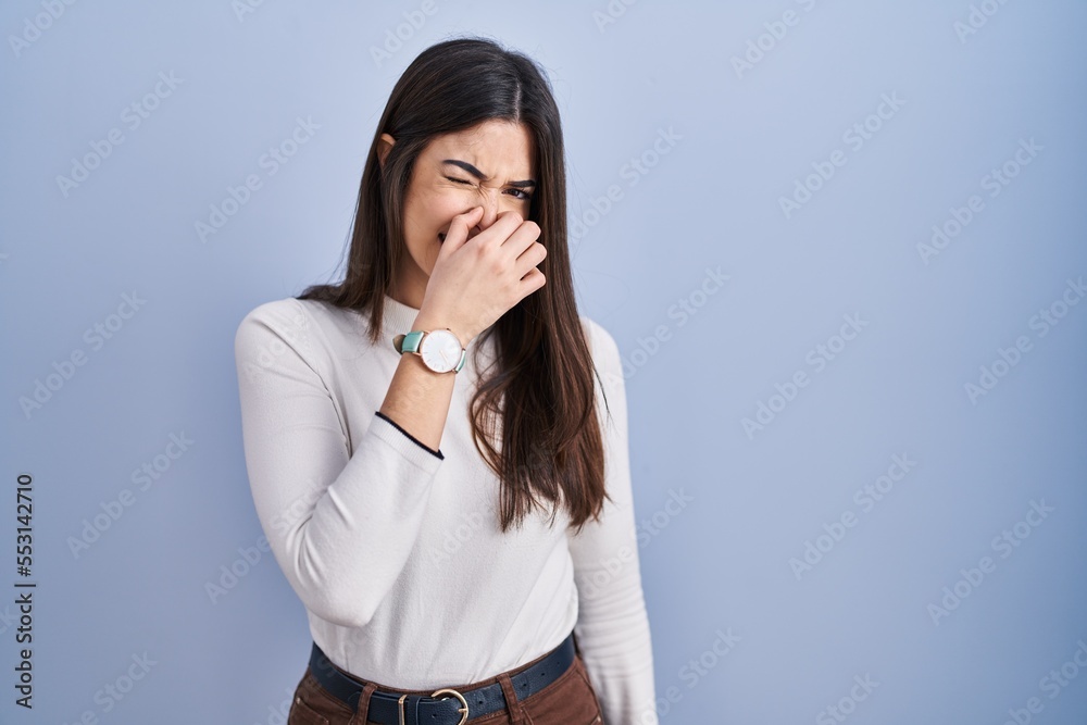 Young brunette woman standing over blue background smelling something ...