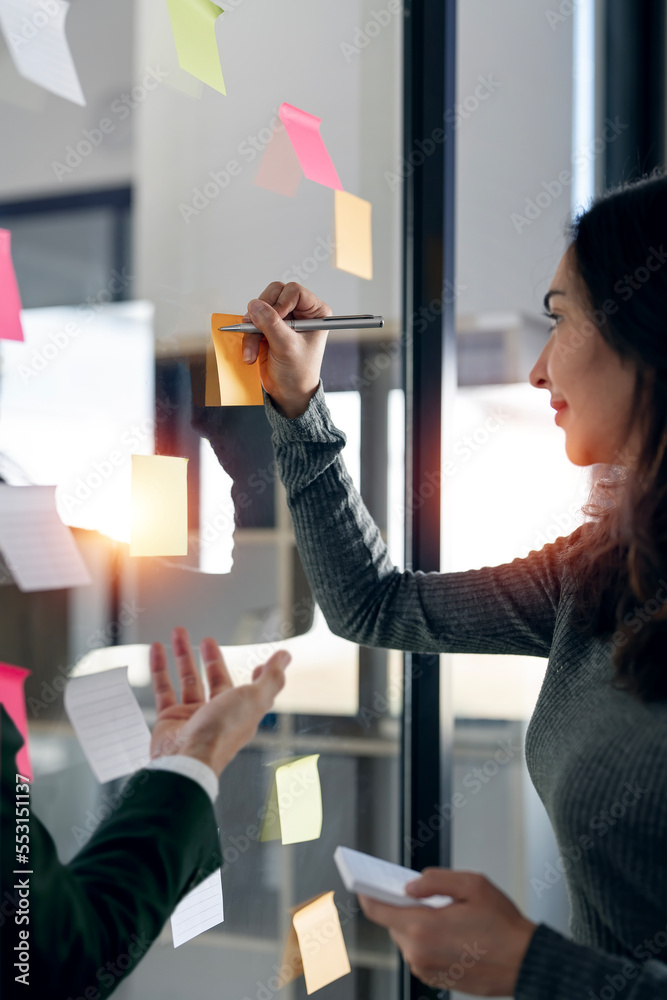 Two young creative team using post it notes in glass wall to writing ...