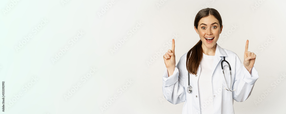 Enthusiastic young woman doctor smiling, pointing fingers up, wearing ...