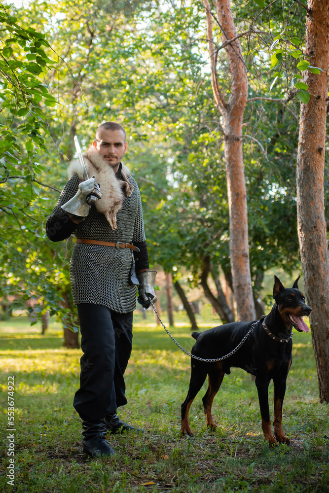 A young man in chain mail with a sword in the woods with a dog. A ...