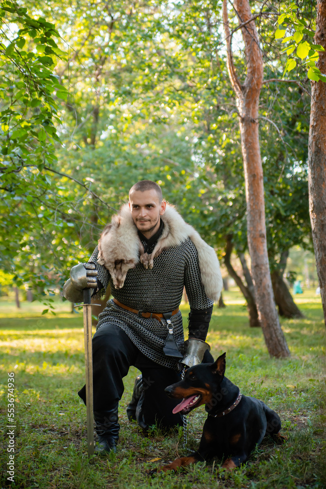 A young man in chain mail with a sword in the woods with a dog. A ...
