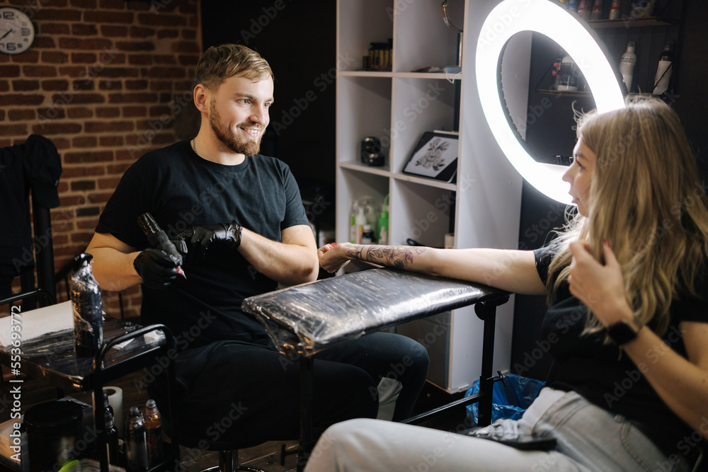 Young smiled tattoo artist is tattooing a woman's hand. Wireless tattoo ...