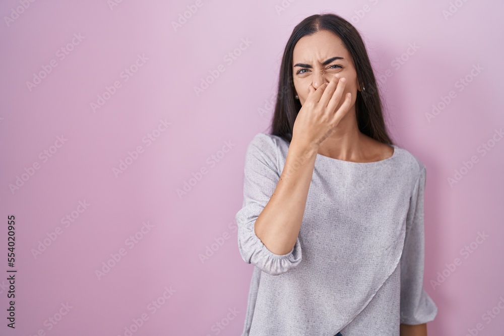 Young brunette woman standing over pink background smelling something ...