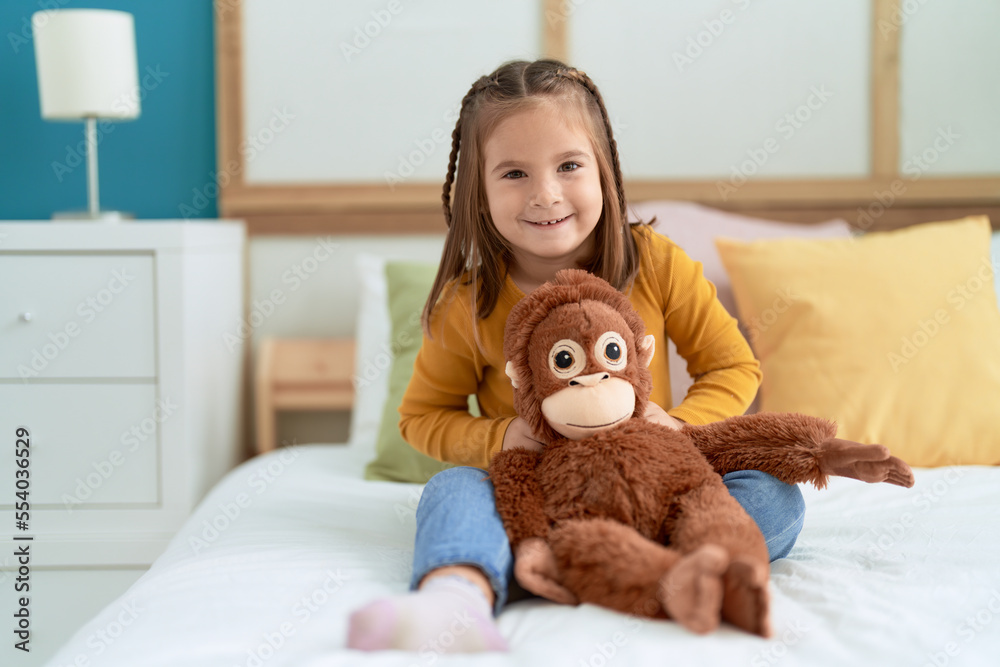 Adorable hispanic girl hugging monkey doll sitting on bed at bedroom ...