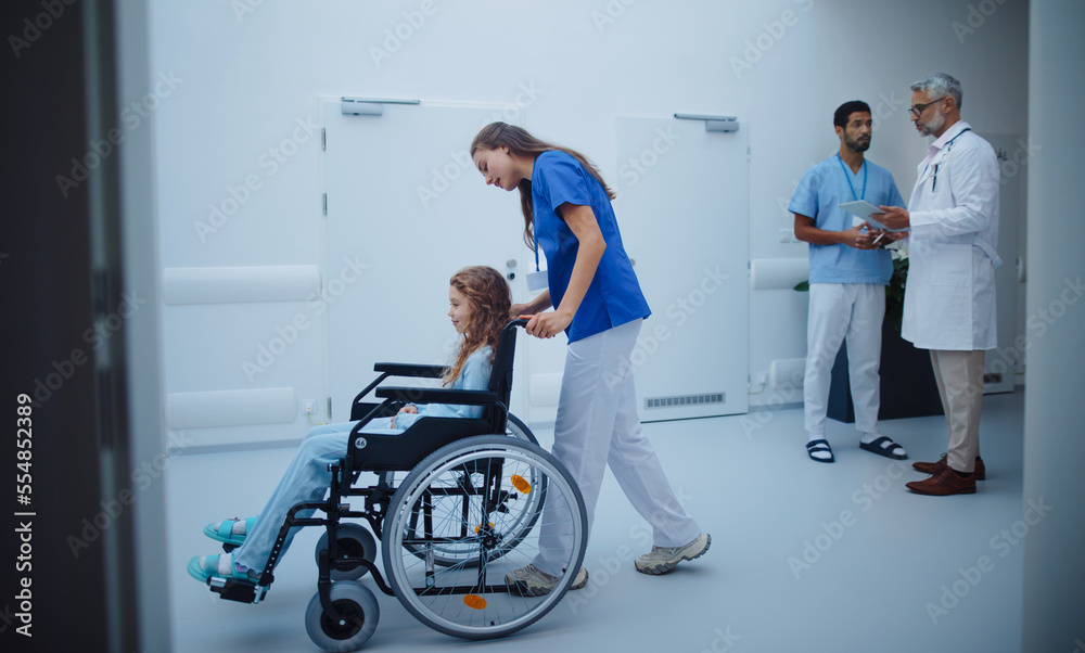 Young nurse pushing little girl on wheelchair at hospital corridor ...